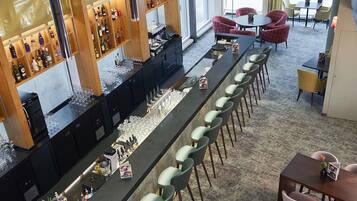Uma área de bar com um balcão extenso, diversas opções de assento e vista da cidade através de amplas janelas.