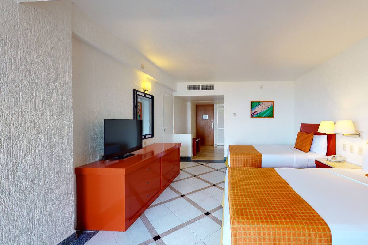 Quarto de hotel com televisão sobre um móvel de madeira, cama com roupa de cama laranja, mesa de cabeceira com abajur e um quadro na parede.