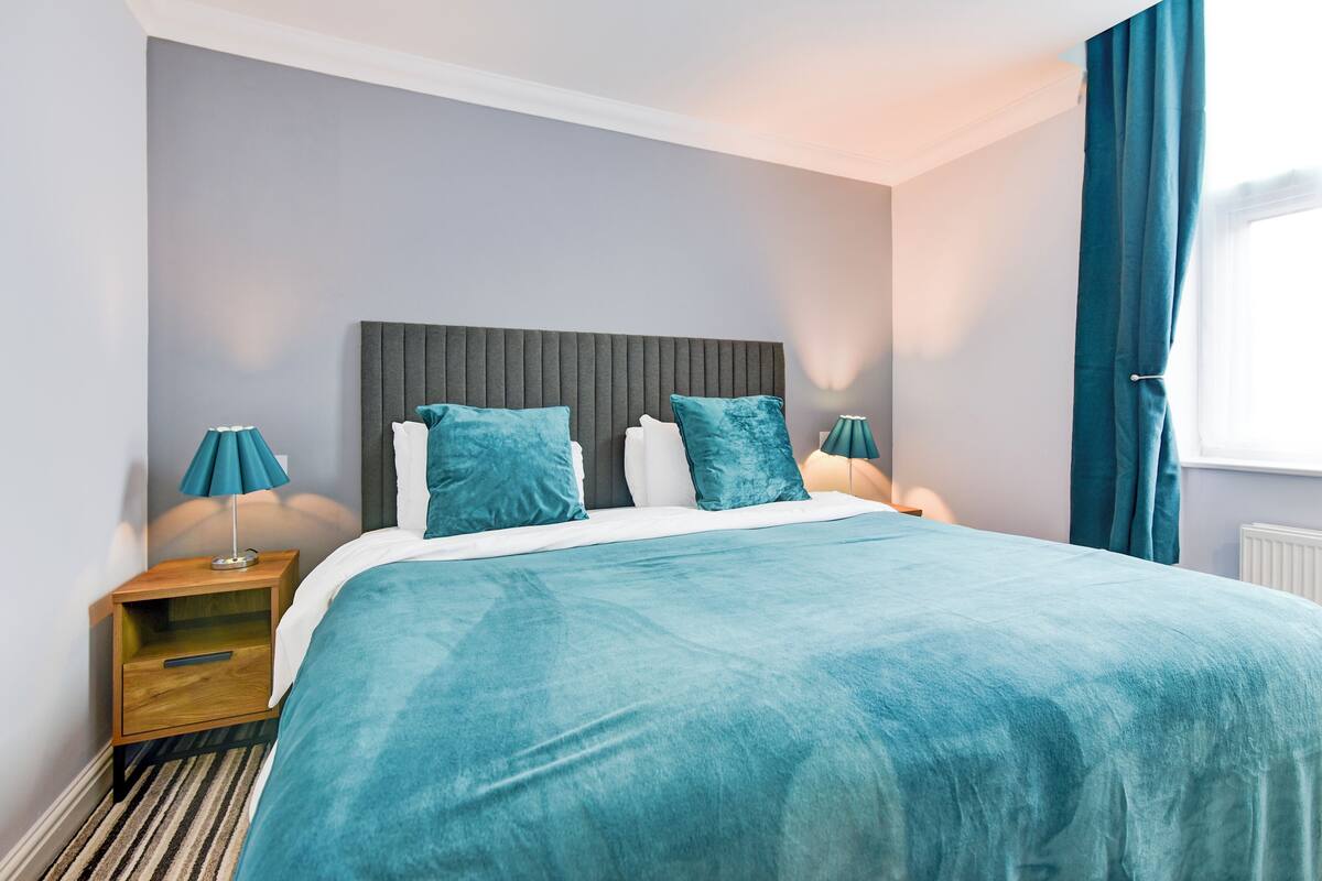 Uma cama bem arrumada com colcha e almofadas verde-água, uma mesa de cabeceira de madeira com abajur e uma janela com cortinas azuis.