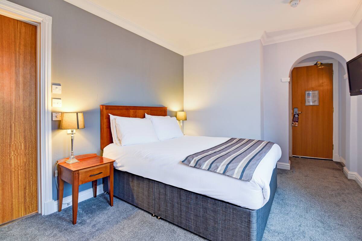Uma cama bem arrumada com cabeceira de madeira, mesa de cabeceira com abajur e piso coberto por carpete.