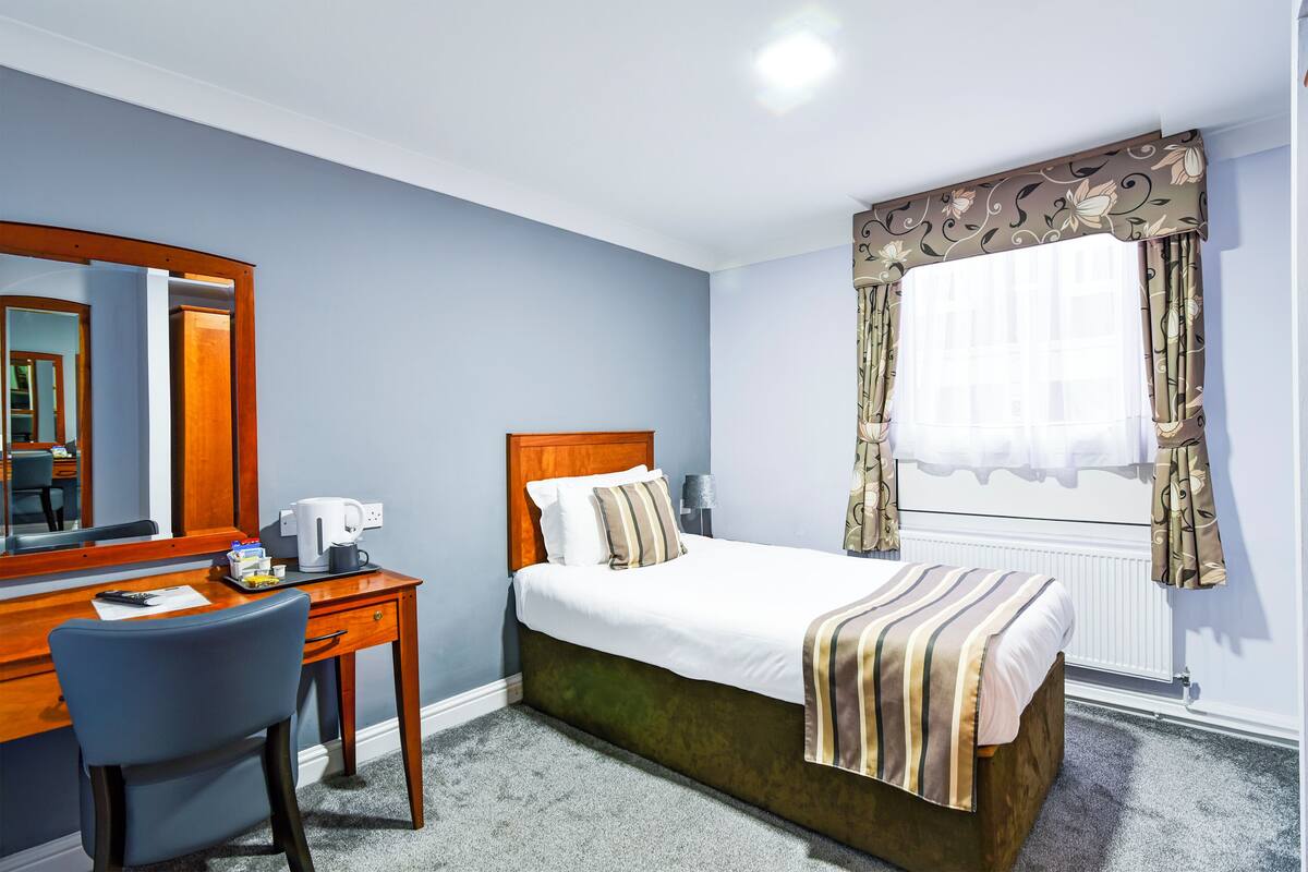 Quarto de hotel com uma cama, uma escrivaninha com cadeira, um espelho e uma janela com cortinas.