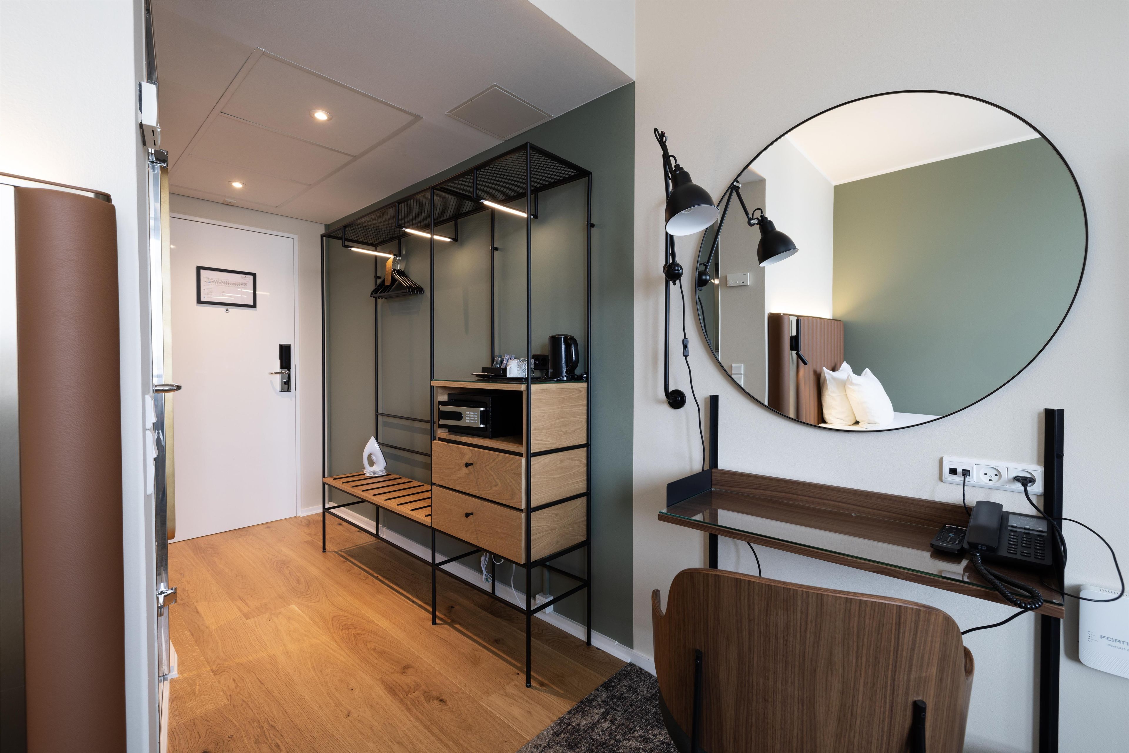 Quarto de hotel com piso de madeira, uma escrivaninha com cadeira, espelho e guarda-roupa com cabideiro.
