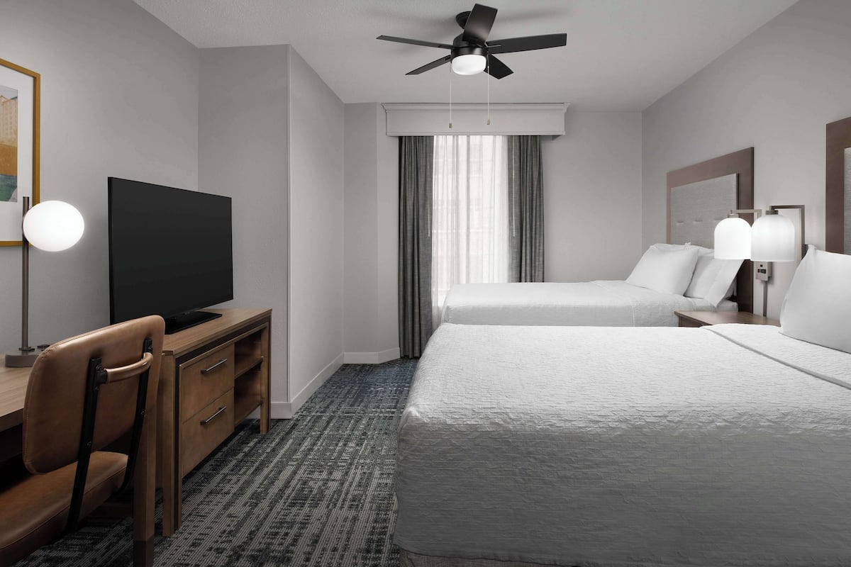 Quarto de hotel com cama, escrivaninha com televisão, cadeira, ventoinha no teto e janela com cortinas.