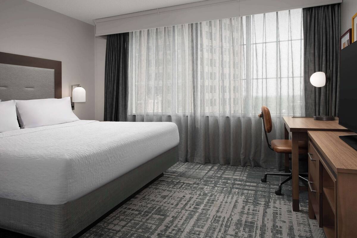 Quarto de hotel com uma cama grande, uma escrivaninha, uma cadeira e uma janela com cortinas.