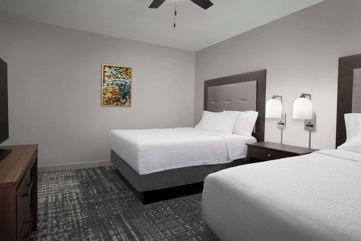 Quarto de hotel com duas camas, uma escrivaninha, televisão, ventilador de teto e um quadro na parede.