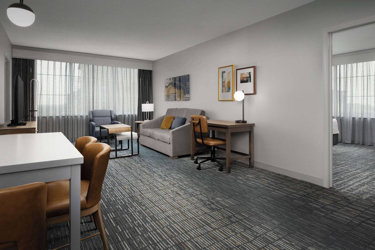Quarto de hotel moderno com sofá cinzento, escrivaninha de madeira, mesa de jantar com cadeiras e uma janela ampla com cortinas leves.