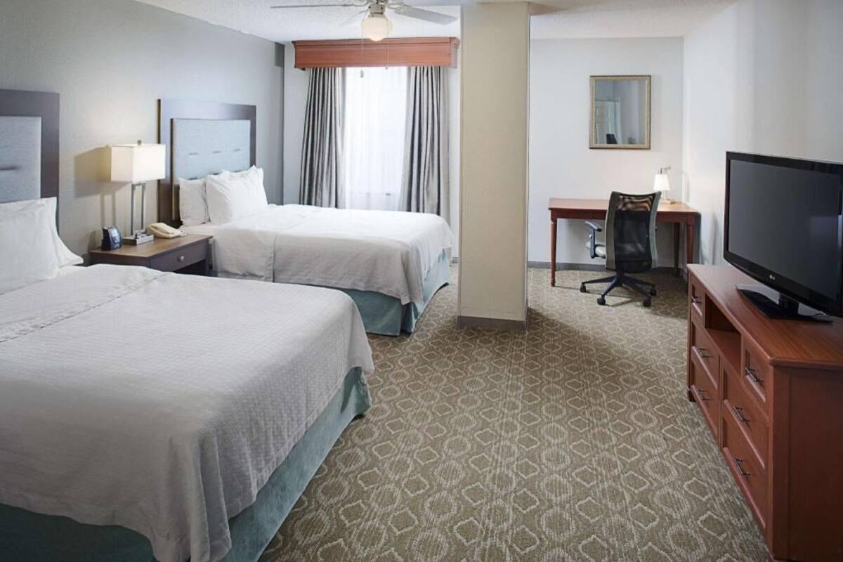 Quarto de hotel com duas camas, uma escrivaninha com cadeira, televisão e ventoinha no teto.