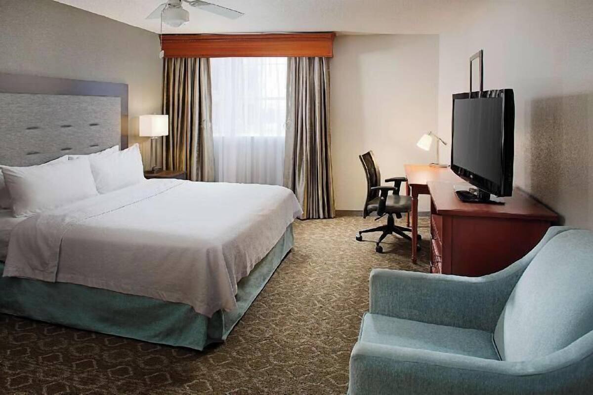 Quarto de hotel com cama, escrivaninha com televisão, cadeira e janela com cortinas.