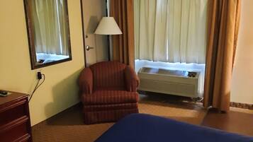 Quarto de hotel com cama, poltrona, escrivaninha, luminária e janela com cortinas.