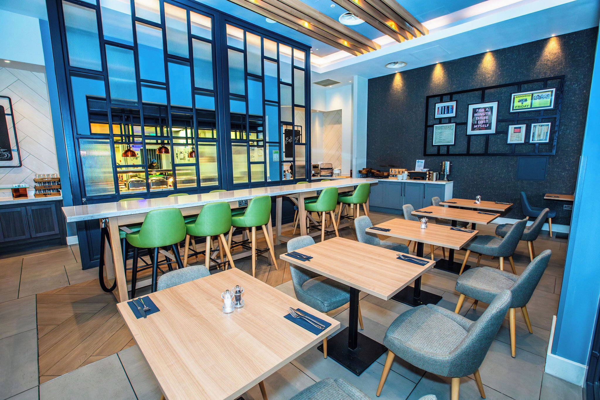 Um restaurante moderno com mesas de madeira e cadeiras verdes, cozinha visível através de amplas janelas e quadros decorativos na parede.