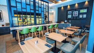 Um restaurante moderno com mesas de madeira e cadeiras verdes, cozinha visível através de amplas janelas e quadros decorativos na parede.