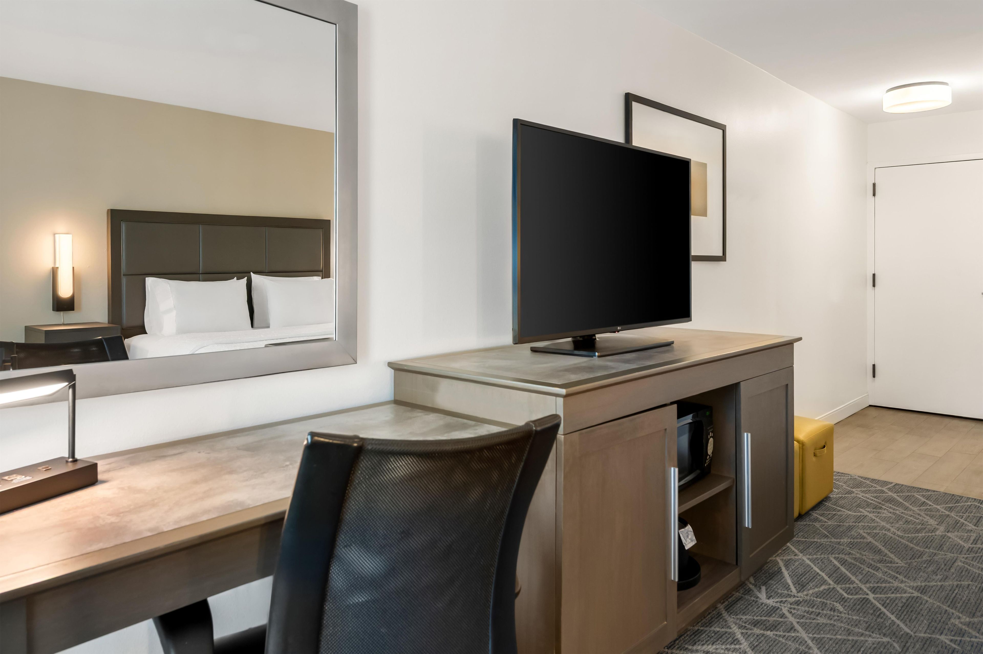 Quarto de hotel com cama, escrivaninha, cadeira, televisão e micro-ondas.