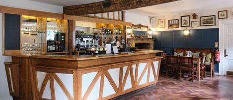 Uma área de bar com balcão de madeira, copos, garrafas e uma máquina de café. Há também um espaço com cadeiras e uma mesa.