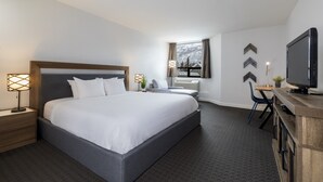 Quarto de hotel moderno com uma cama grande, mesas de cabeceira com luminárias, uma escrivaninha com cadeira, televisão e uma janela com vista para uma paisagem nevada.