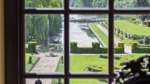 Vista através de uma janela de um jardim bem cuidado, com um caminho, um corpo d'água e uma ponte.