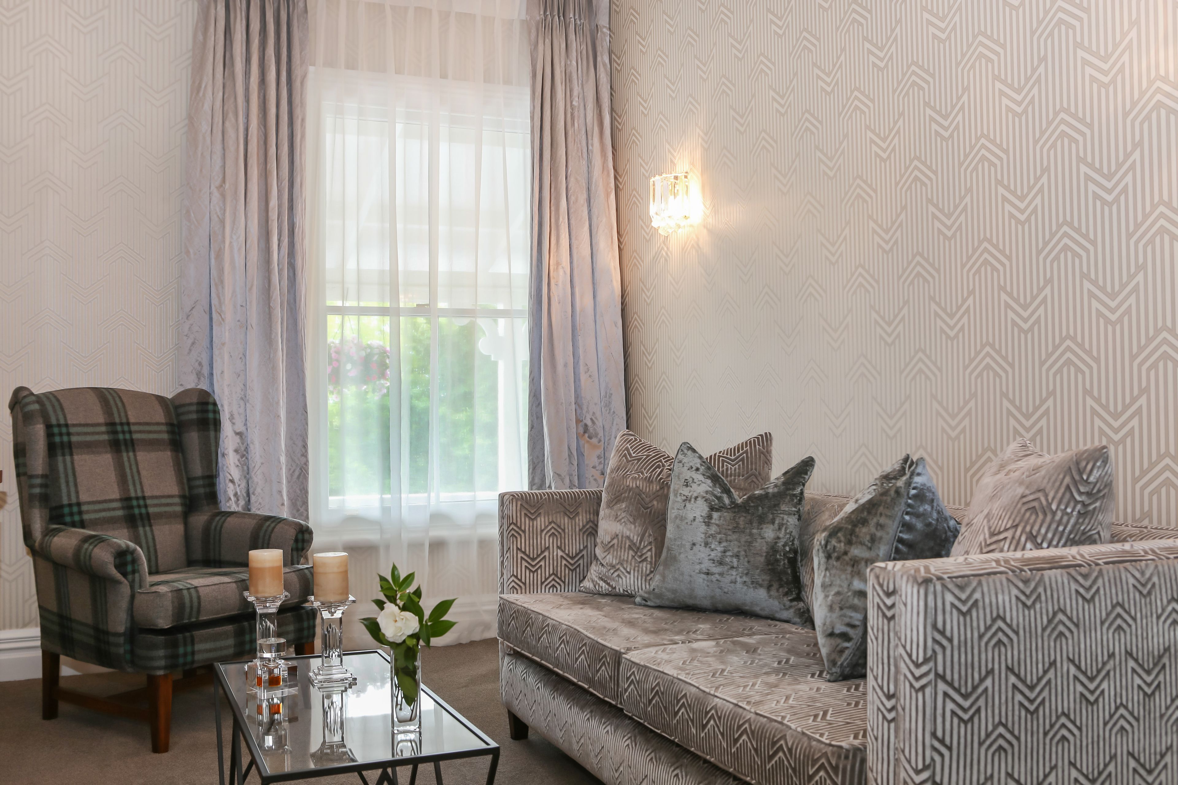 Sala de estar com poltrona xadrez, sofá estampado e mesa de centro de vidro com velas e um vaso.
