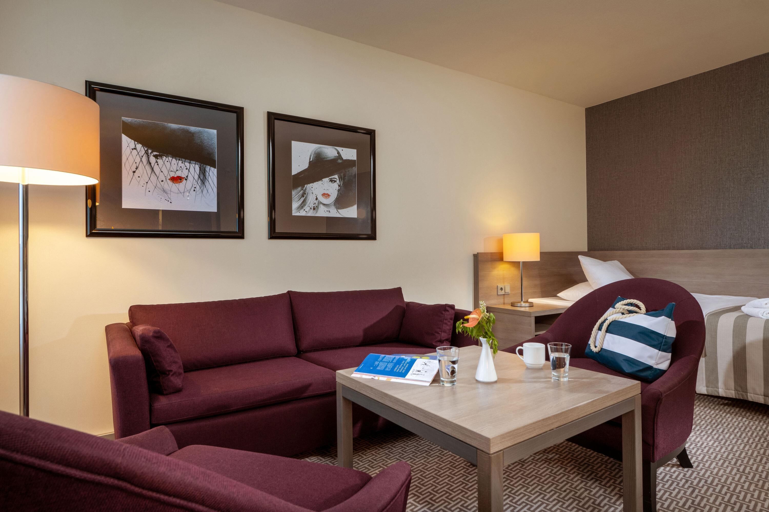 Quarto de hotel com sofá, uma pequena mesa com um livro, um vaso com flores, uma luminária e uma cama com travesseiro e uma sacola listrada.