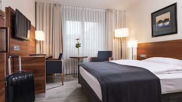 Quarto de hotel com cama, escrivaninha, cadeira, televisão, luminária e mala.