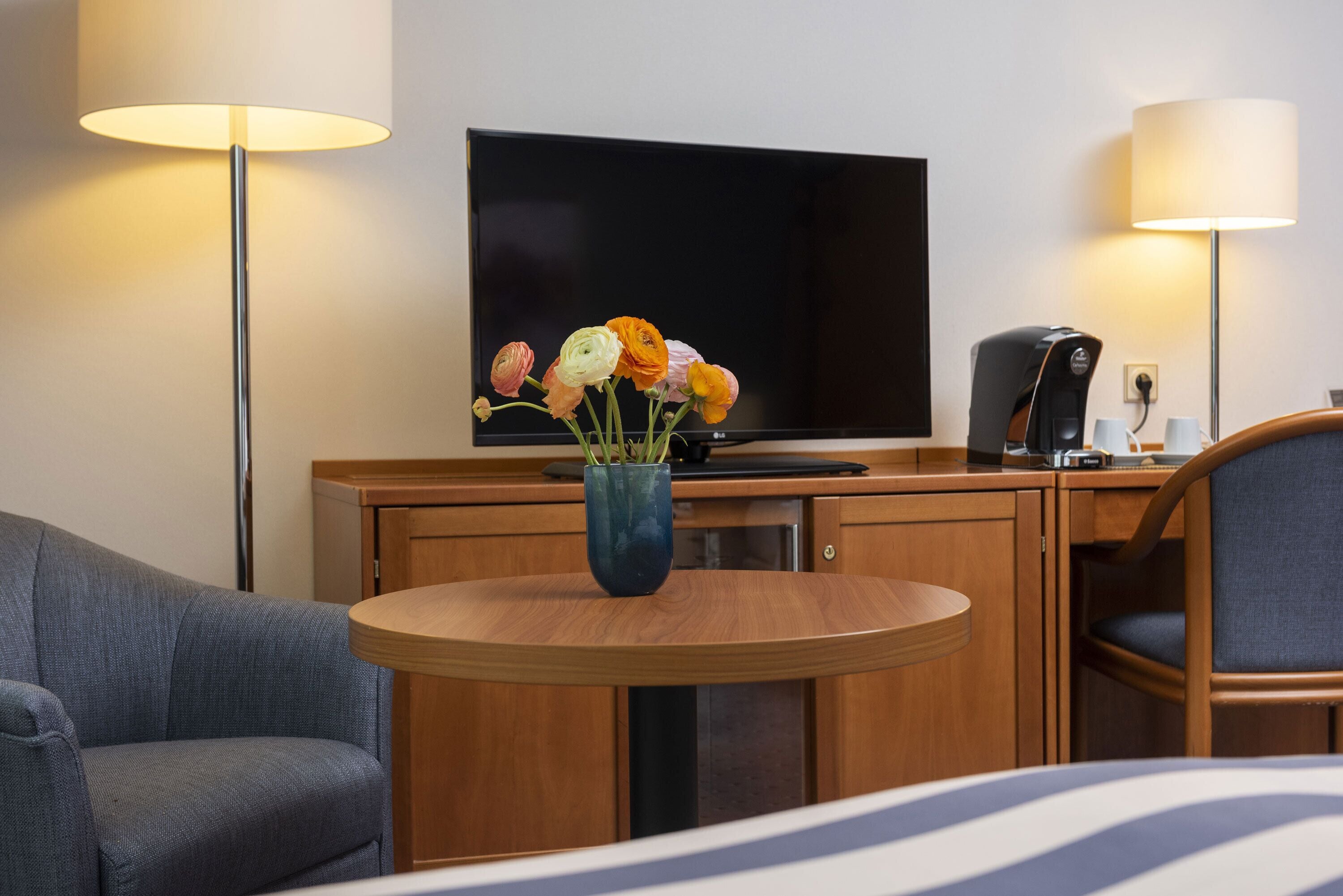 Quarto de hotel com uma mesa redonda de madeira, um vaso azul com flores, uma televisão de tela plana, uma cafeteira e um sofá.