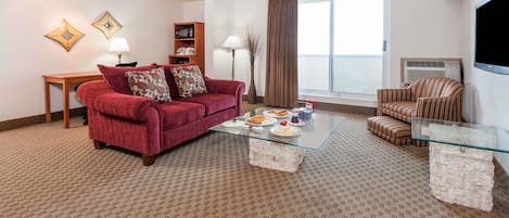 Uma sala de estar com um sofá vermelho, uma mesa de centro de vidro e uma poltrona listrada. Há uma estante, um abajur e uma janela grande com cortinas.