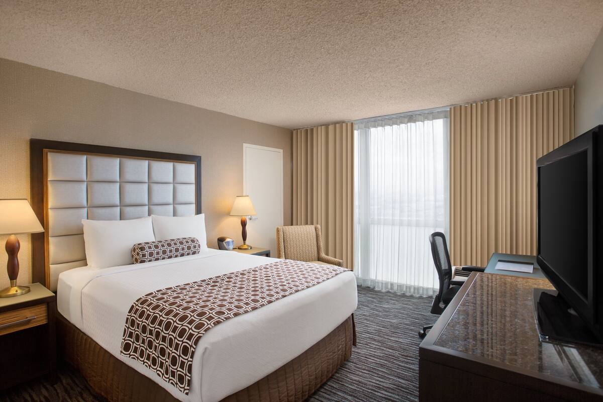 Quarto de hotel com uma cama grande, uma escrivaninha com televisão, uma cadeira e uma janela com cortinas.