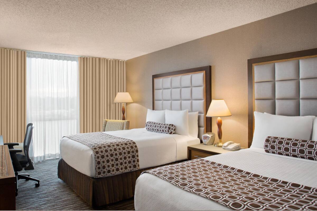 Quarto de hotel com duas camas, uma escrivaninha, uma cadeira, televisão e janela com cortinas.
