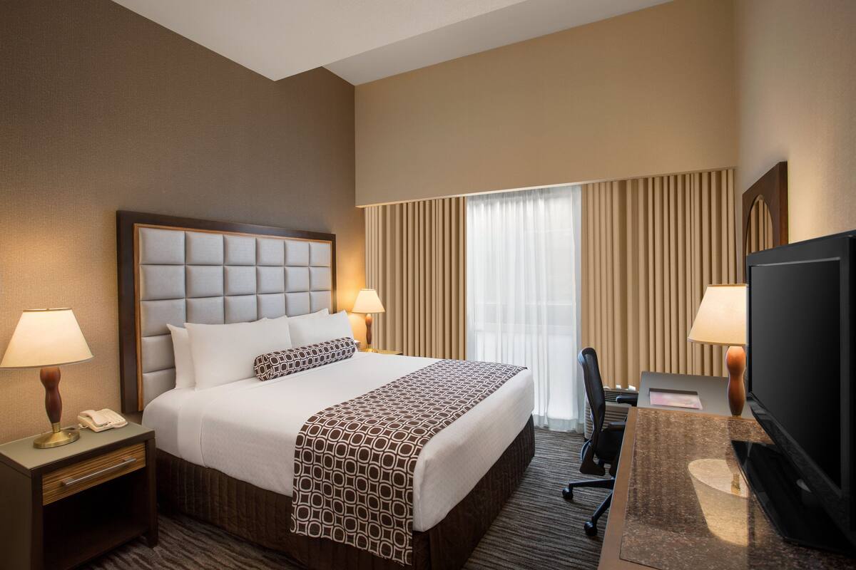 Quarto de hotel com uma cama grande, duas luminárias de cabeceira, uma mesa com televisão e uma janela com cortinas.