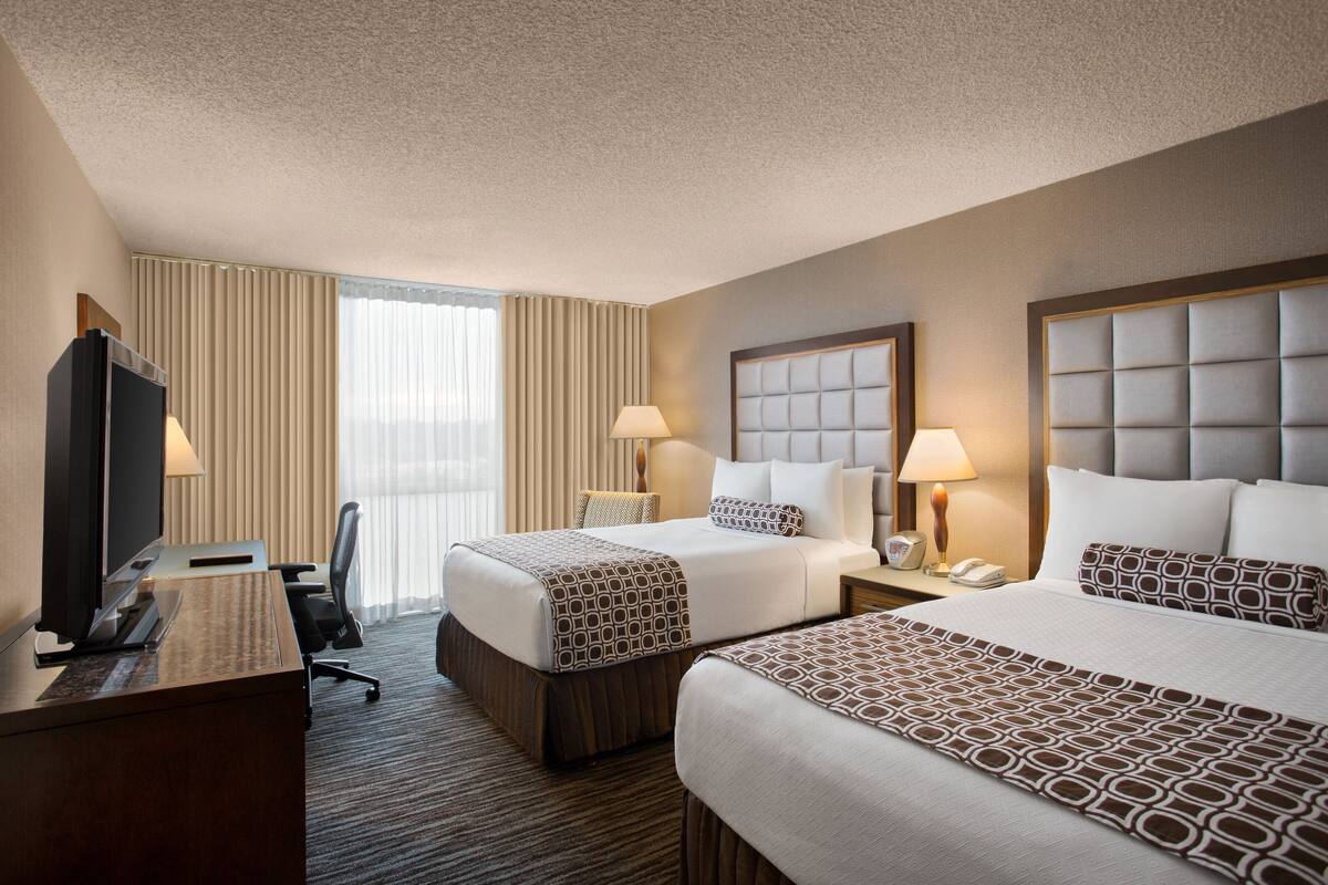 Quarto de hotel com duas camas, uma escrivaninha, uma televisão e uma janela com cortinas.