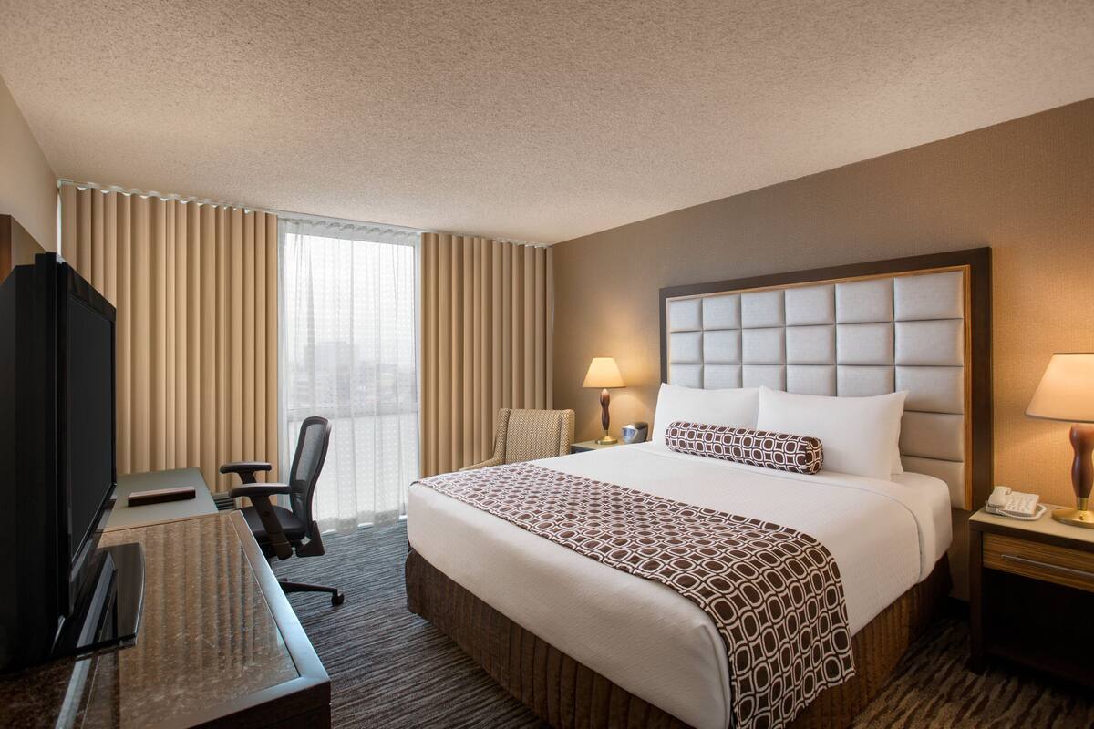 Quarto de hotel com uma cama grande, uma escrivaninha com cadeira, televisão e janela com cortinas.