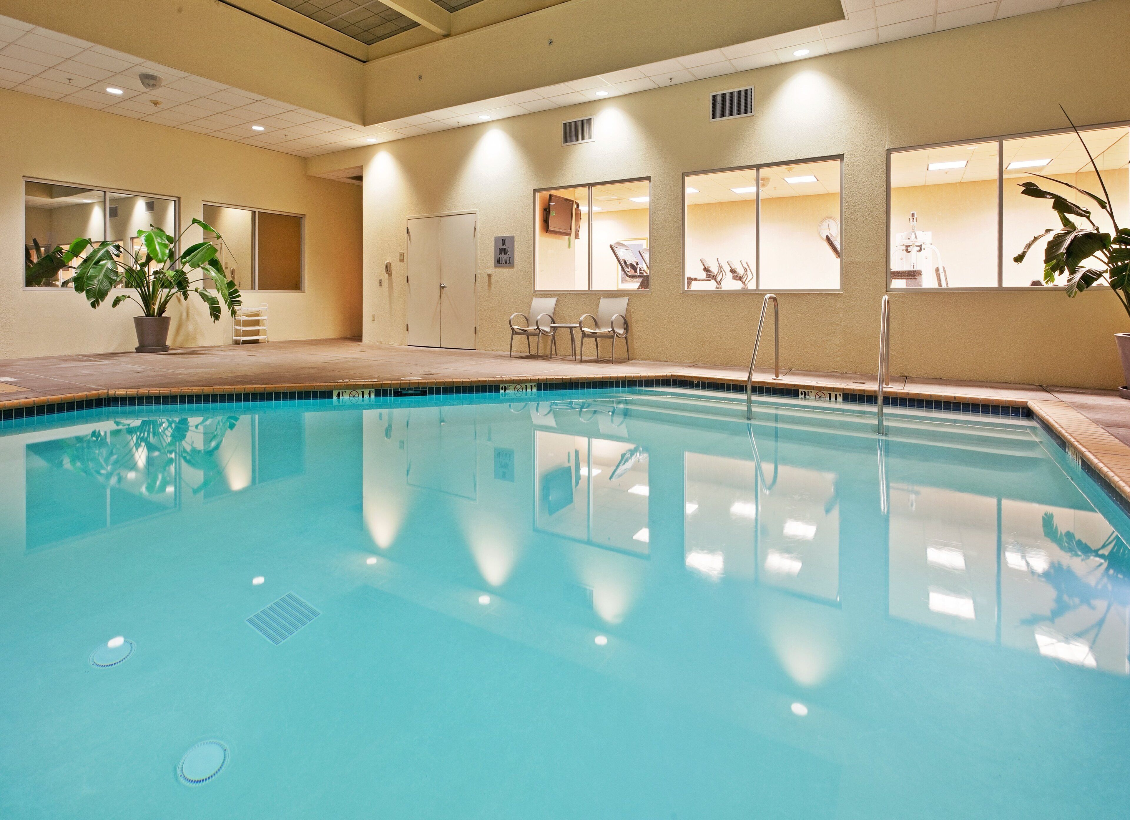 Uma piscina interior com água cristalina, rodeada por paredes com janelas e portas, e um chão ladrilhado.