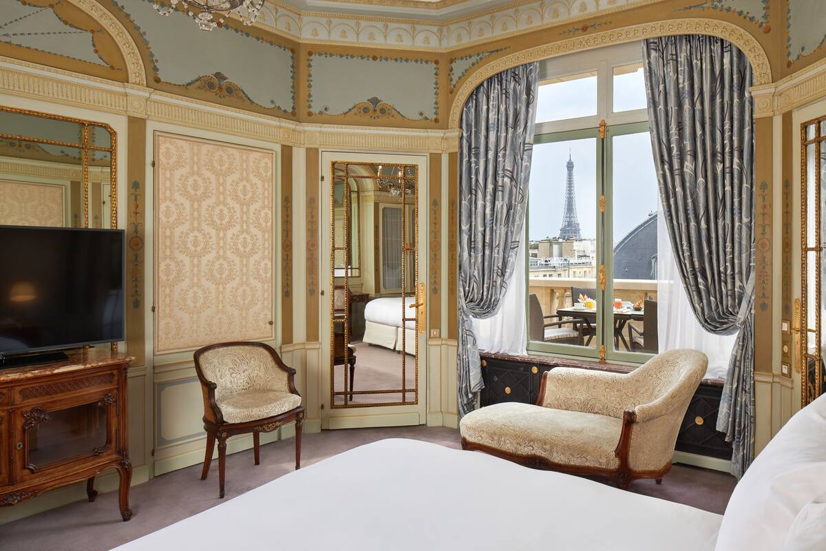 Quarto de hotel luxuoso com uma cama grande, televisão, duas poltronas e vista para a Torre Eiffel.