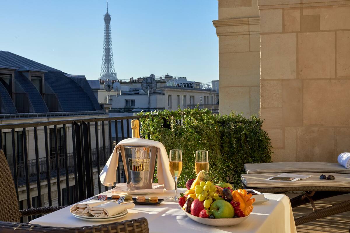 Varanda com mesa posta para uma refeição, uma garrafa de champanhe e vista para a Torre Eiffel.