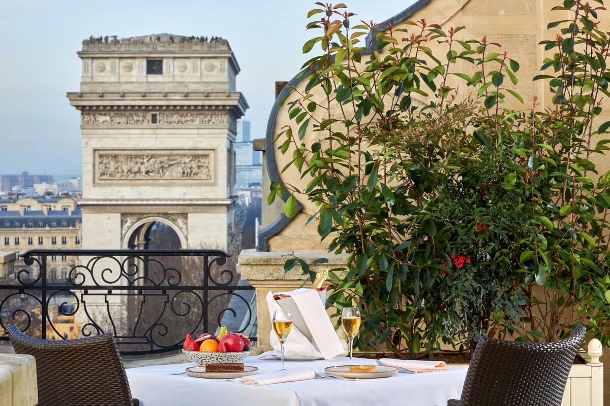 Terraço com vista para um monumento histórico, cadeiras de vime, uma mesa posta com uma cesta de frutas, copos de vinho e um cardápio.