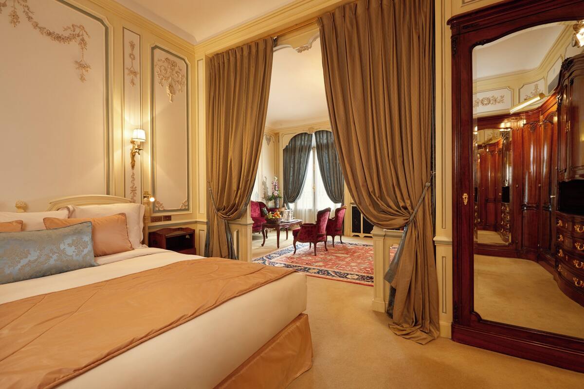 Quarto de hotel com uma cama grande, área de estar com mesa e cadeiras, e um espelho.