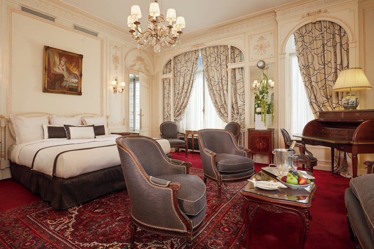 Quarto de hotel luxuoso com uma cama grande, duas poltronas, uma mesa de jantar com um prato de frutas e um piano.