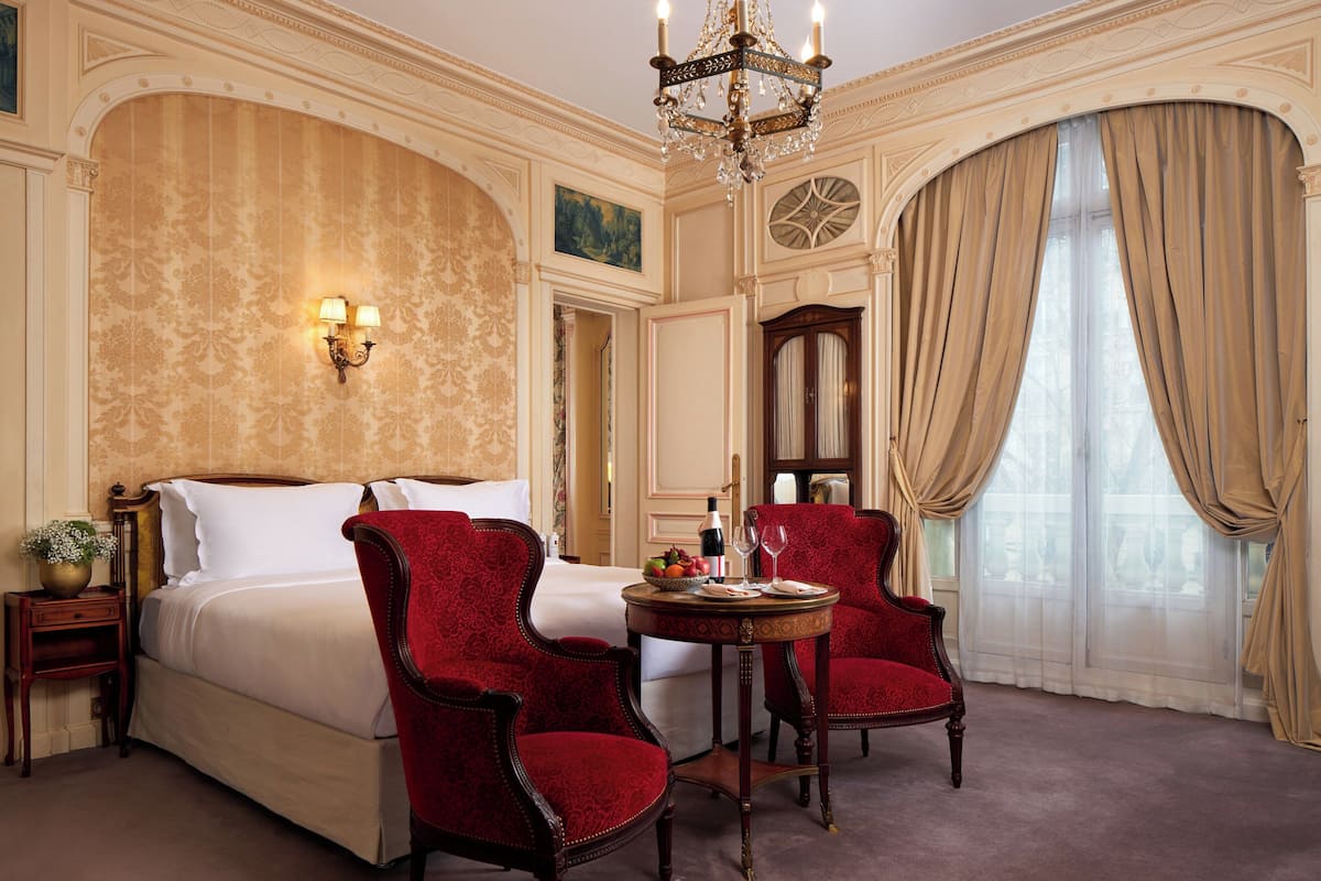 Quarto de hotel luxuoso com uma cama grande, duas poltronas vermelhas, uma mesinha redonda com uma taça de vinho e um candelabro.