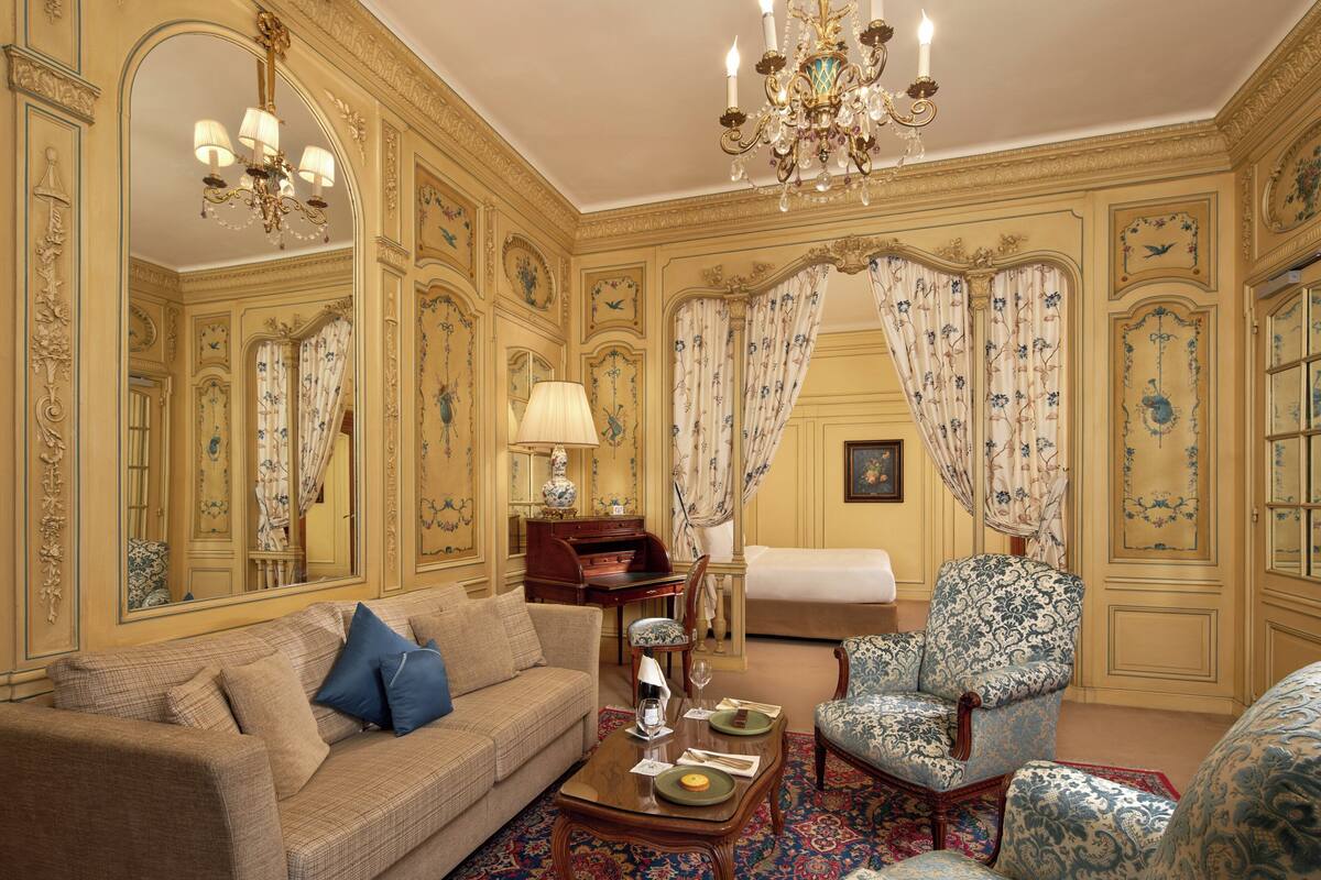 Um quarto ricamente decorado, com um lustre grande e ornamentado, um sofá macio com almofadas azuis, uma poltrona estampada e uma mesa lateral de madeira clássica.