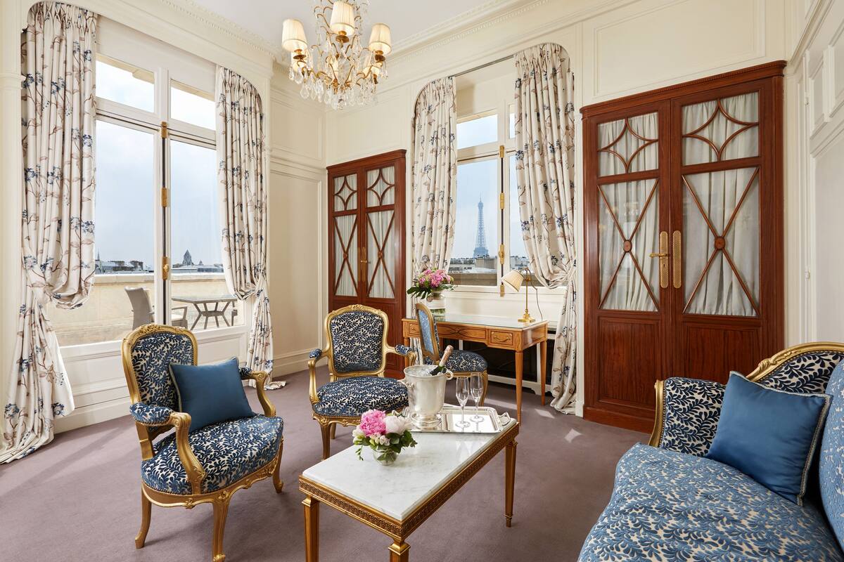 Quarto com duas poltronas azuis estampadas, uma mesa branca com um vaso de flores e vista para a Torre Eiffel através de grandes janelas com cortinas.