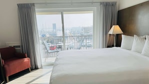 Quarto de hotel com uma cama grande, uma poltrona vermelha, uma varanda com vista para barcos e edifícios, e um abajur na mesa de cabeceira.