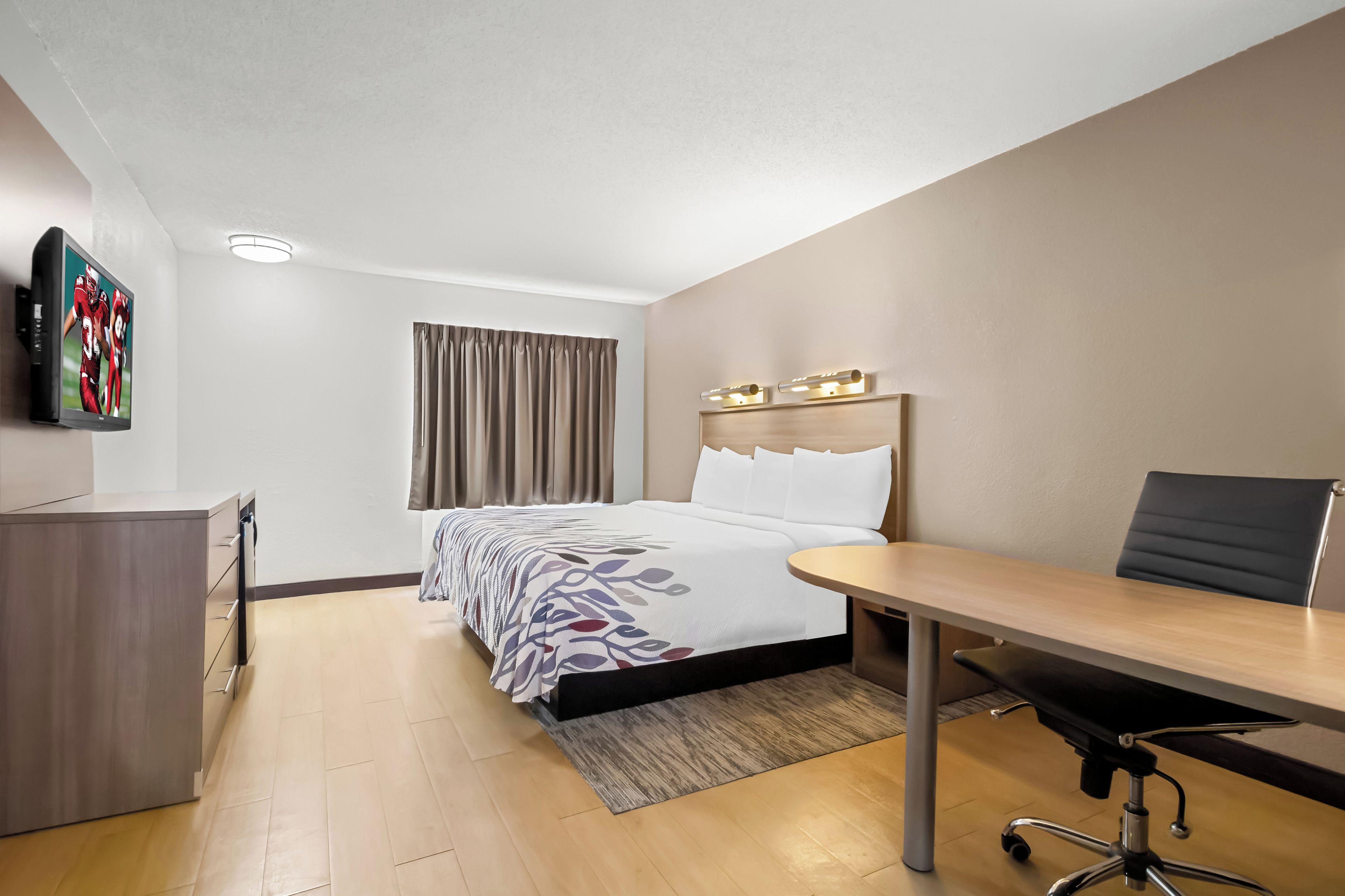 Quarto de hotel com cama, escrivaninha, cadeira e televisão na parede.