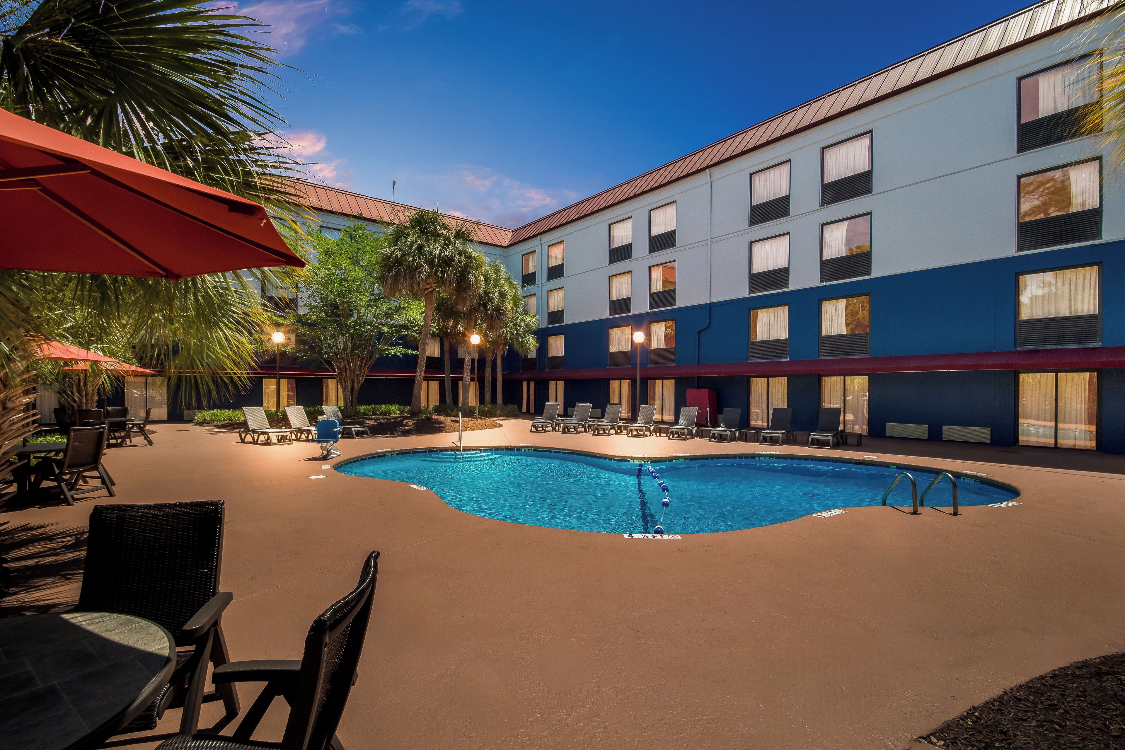Área de piscina de hotel com várias espreguiçadeiras e guarda-sóis, rodeada por um edifício com várias janelas.
