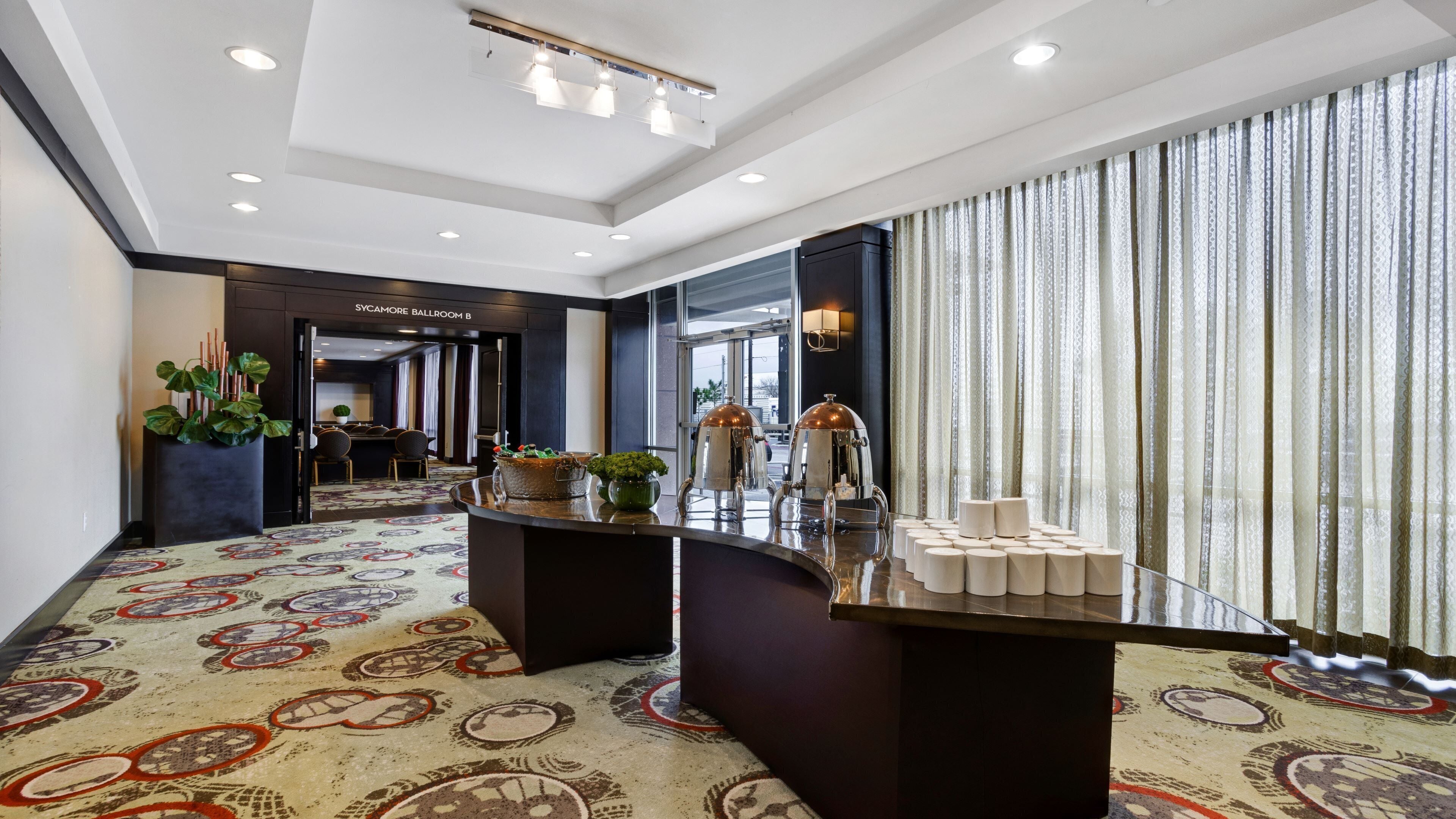 Um hall de hotel com uma mesa de recepção, carpete estampado, uma janela grande com cortinas e uma porta visível que dá para a salão de baile.