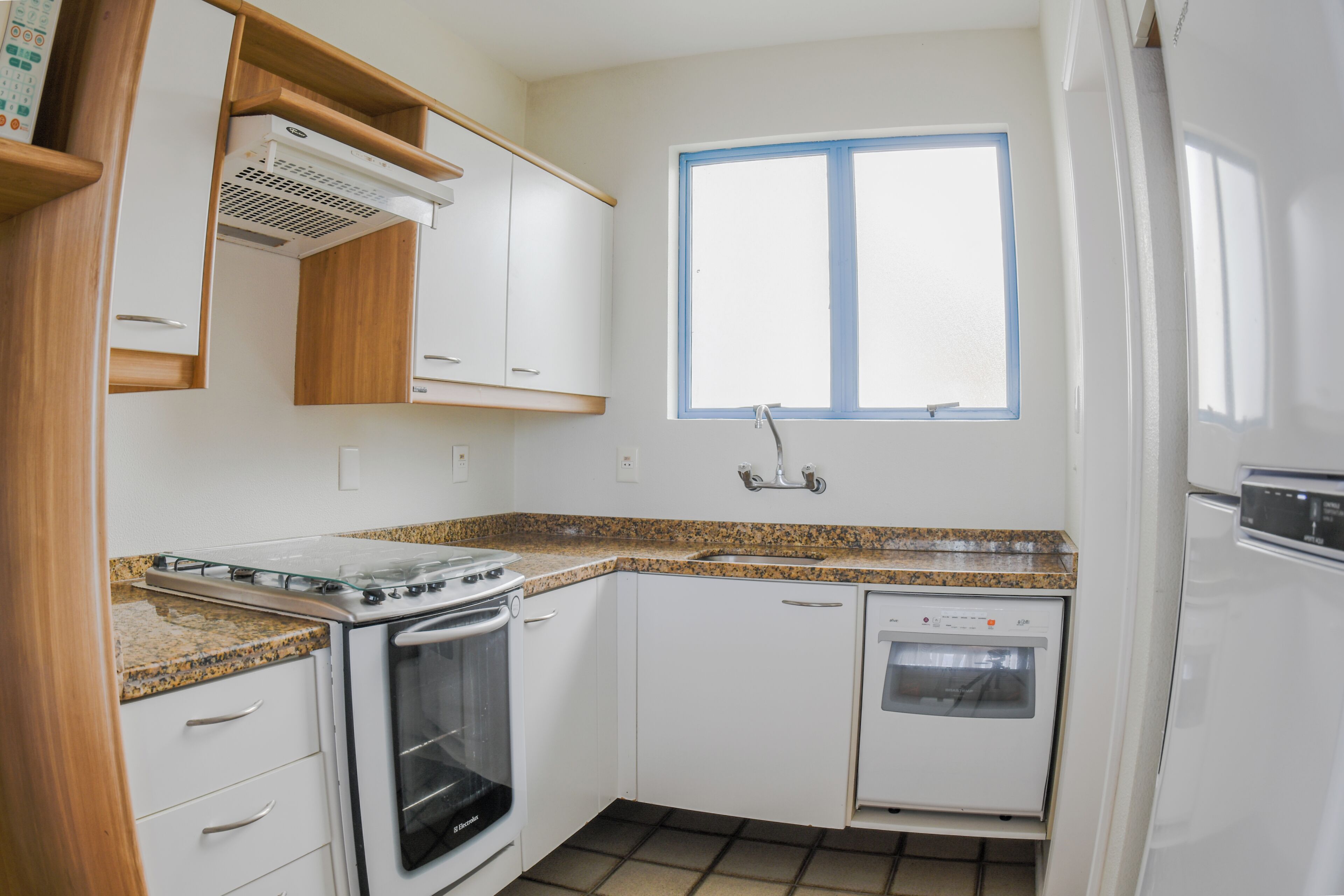 Cozinha compacta com armários brancos, forno embutido, lava-louças e janela com moldura azul.