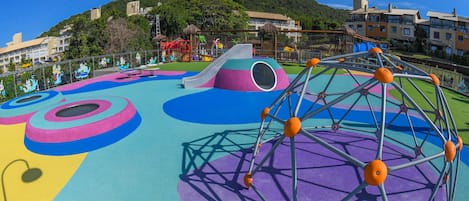 Um parque infantil colorido com um escorrega e uma estrutura de escalada em cúpula, rodeado por edifícios e uma colina.