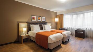Quarto de hotel com duas camas, uma mesinha de cabeceira com abajur, uma cadeira, uma pequena mesa e uma janela grande com cortinas.