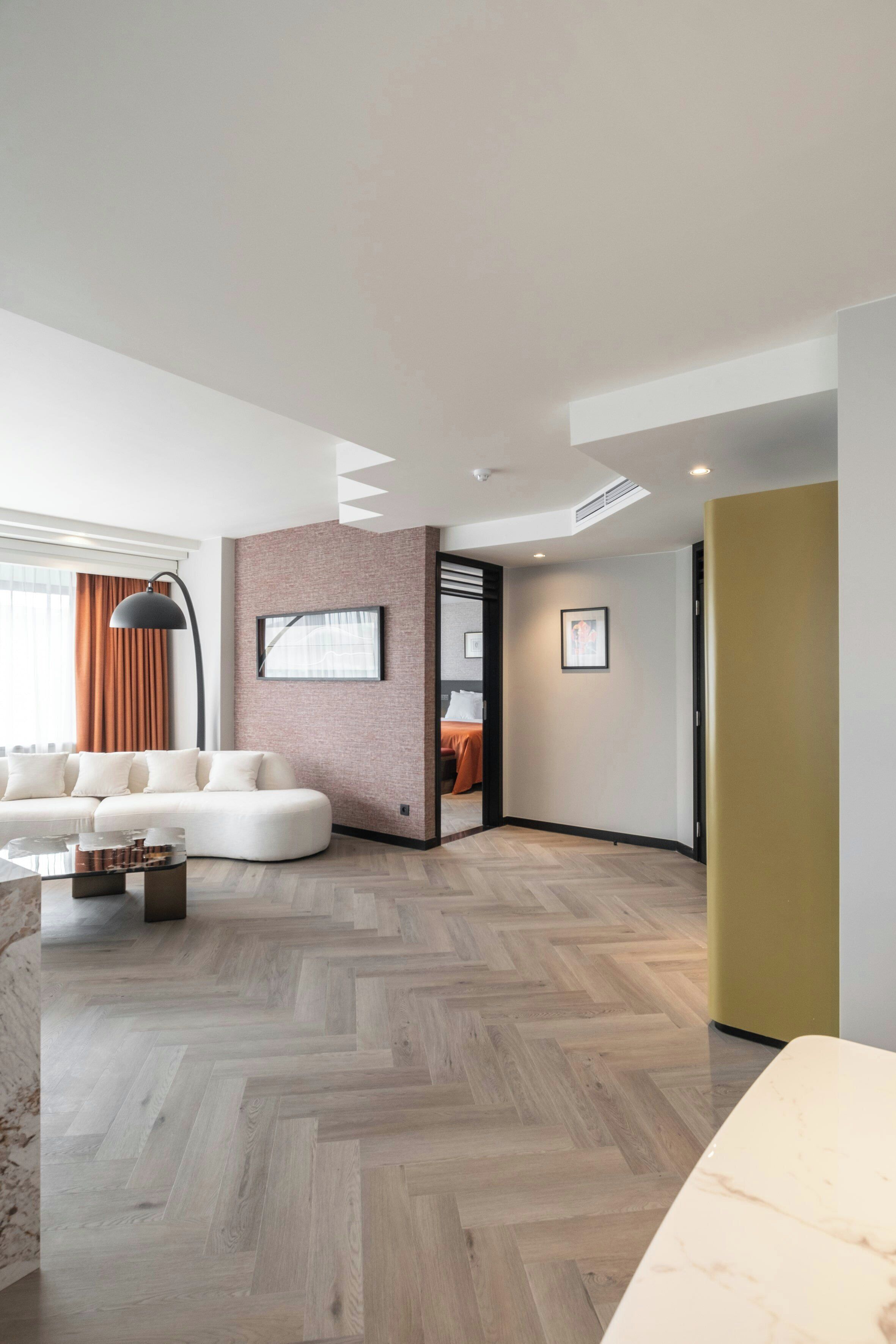 Uma sala de estar moderna com piso de madeira em padrão herringbone, um sofá branco e uma janela grande com cortinas.