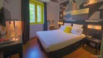 Quarto de hotel com cama, mesas de cabeceira, escrivaninha, janela com cortinas, televisão na parede e um quadro.