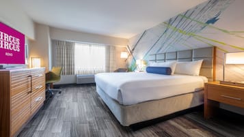 Quarto de hotel com uma cama grande, uma escrivaninha, uma cadeira, um armário e uma janela com cortinas.