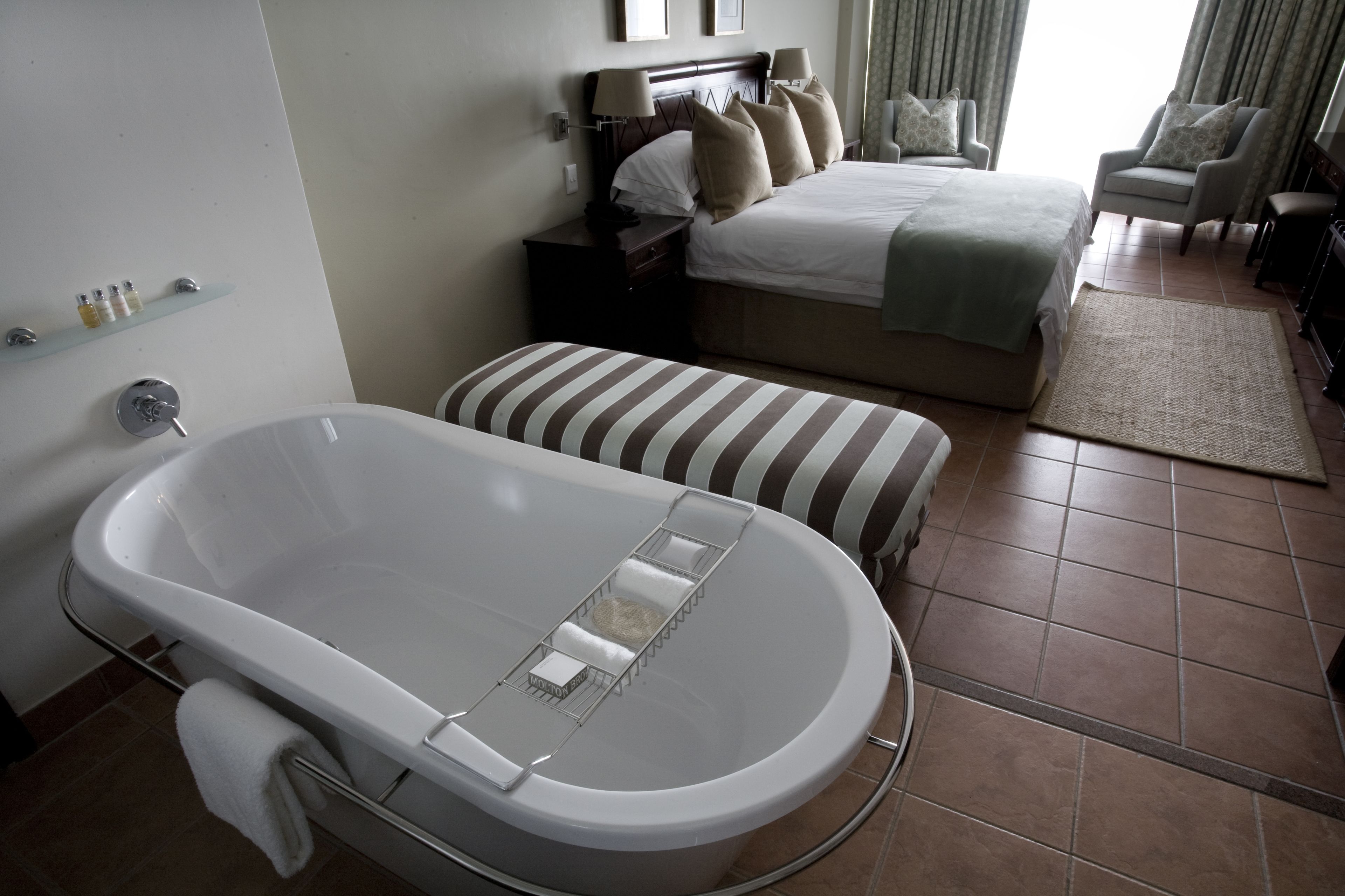 Quarto de hotel moderno com uma banheira grande, uma cama bem arrumada, um espaço de estar com uma poltrona e uma pequena mesa.