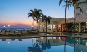 Área da piscina com espreguiçadeiras, um edifício com janelas amplas e vista para o pôr do sol.
